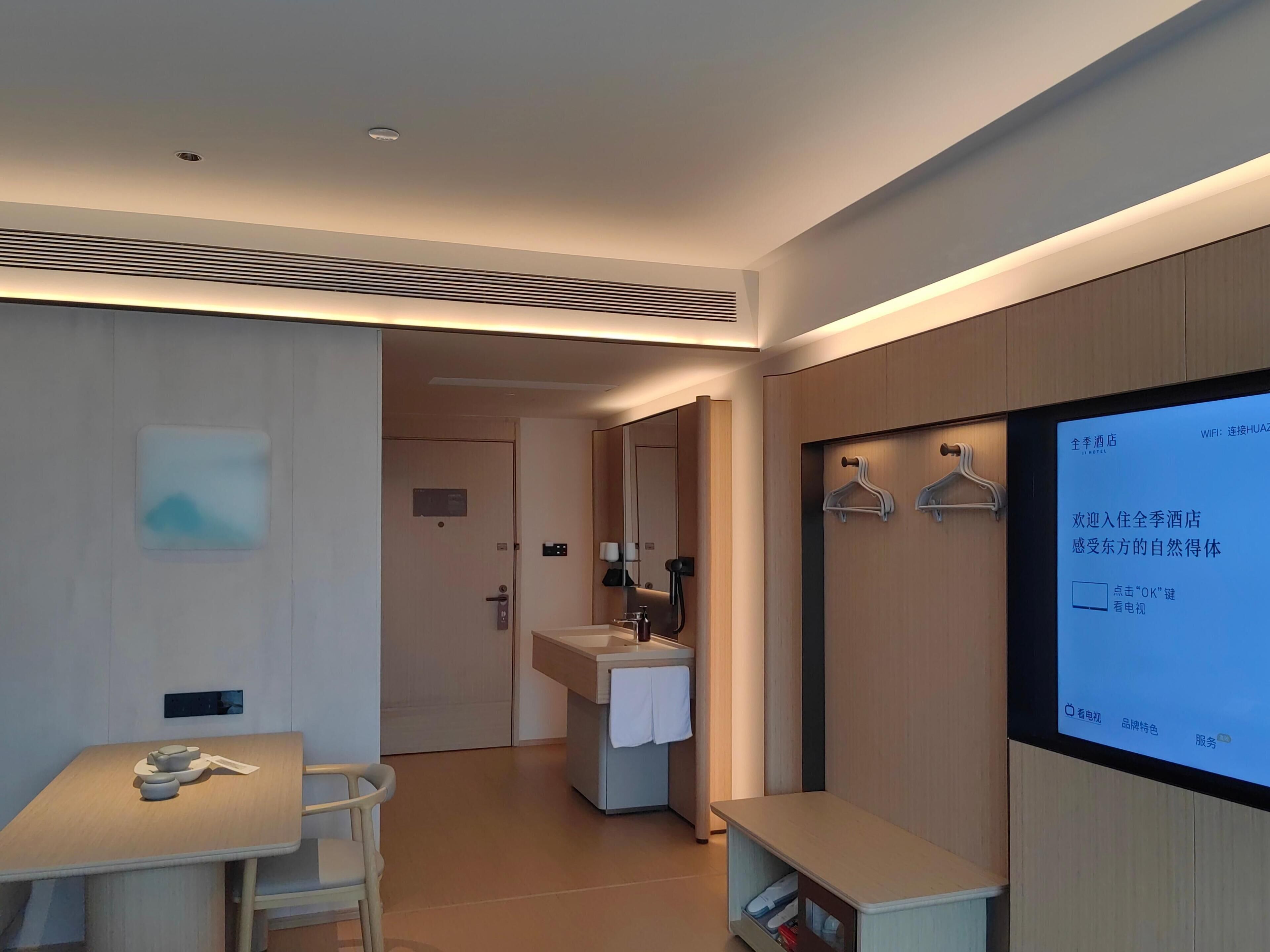Quarto de hotel moderno com televisão de tela plana, escrivaninha com cadeira, banheiro com pia e porta-toalhas, e porta que dá para outro quarto.