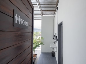 Corredor com parede de madeira, placa indicando um banheiro, uma pia e uma lixeira.
