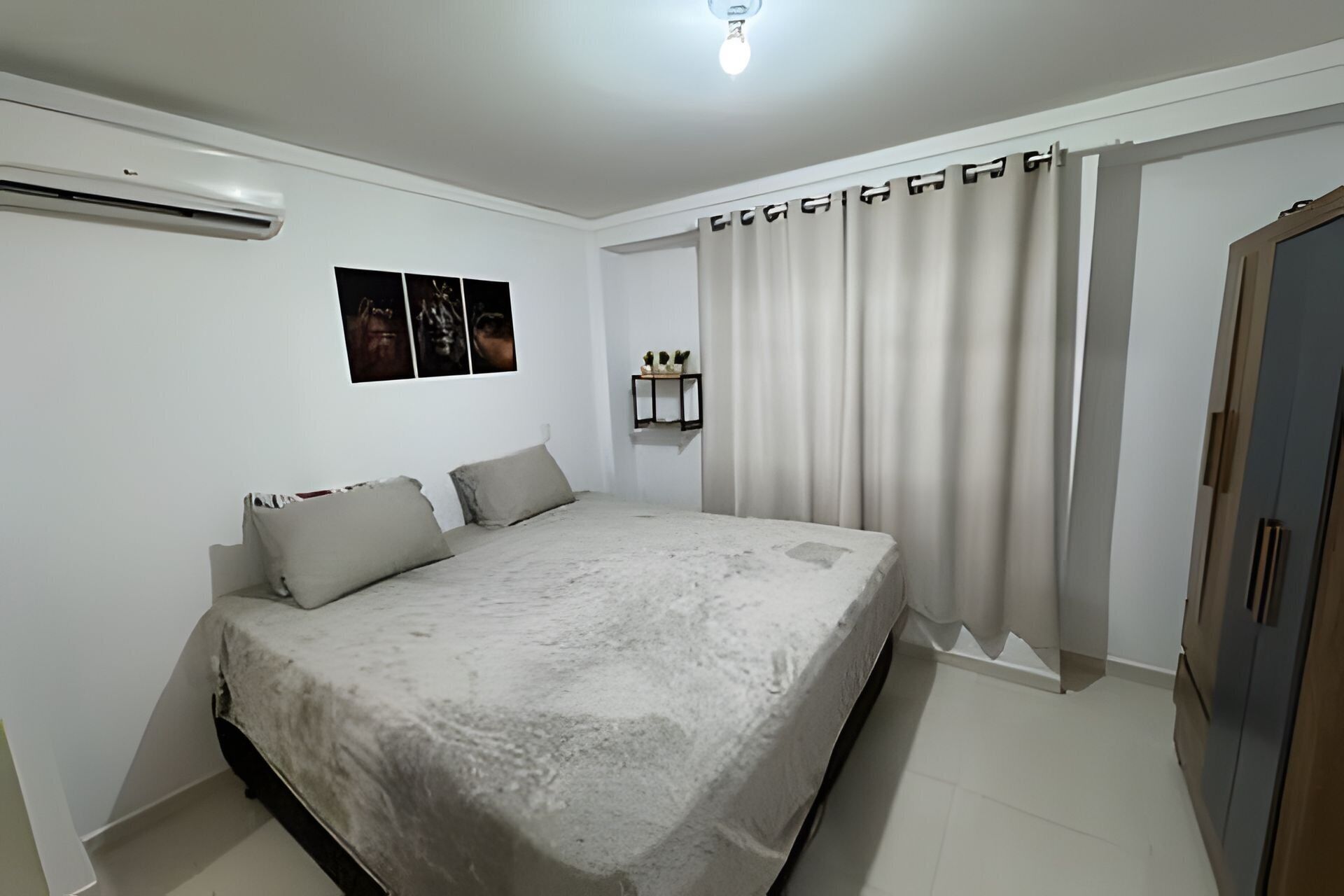Quarto com cama, ar-condicionado na parede, pequeno estante com objetos decorativos e porta.