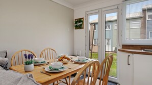 Uma área de refeições com uma mesa de madeira posta para dois, uma pequena planta e uma tigela de comida. Há uma porta que dá para uma varanda com vista para casas.