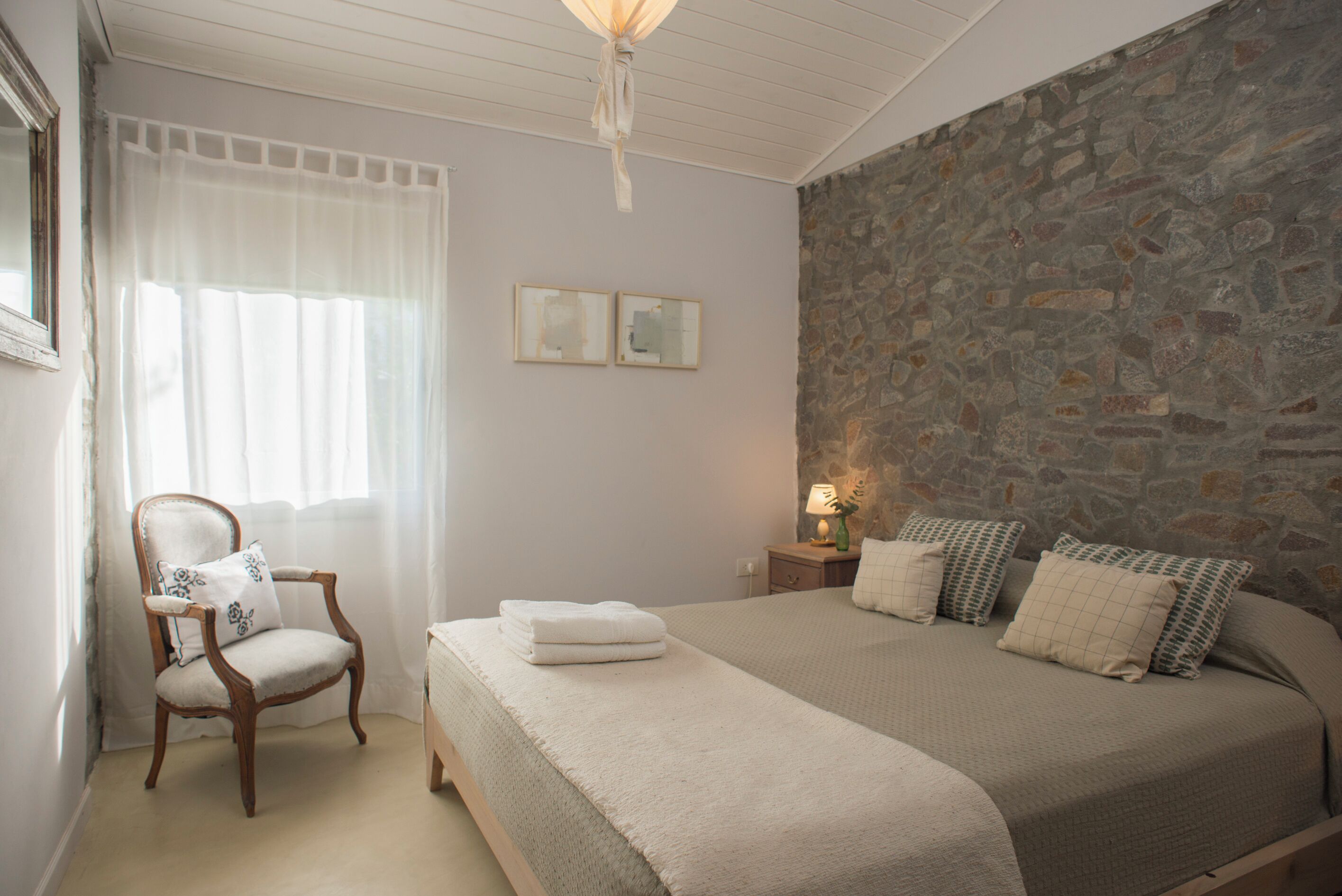 Quarto com parede de pedra, cama com várias almofadas, cadeira com almofada, mesa de cabeceira com abajur e janela com cortinas leves.