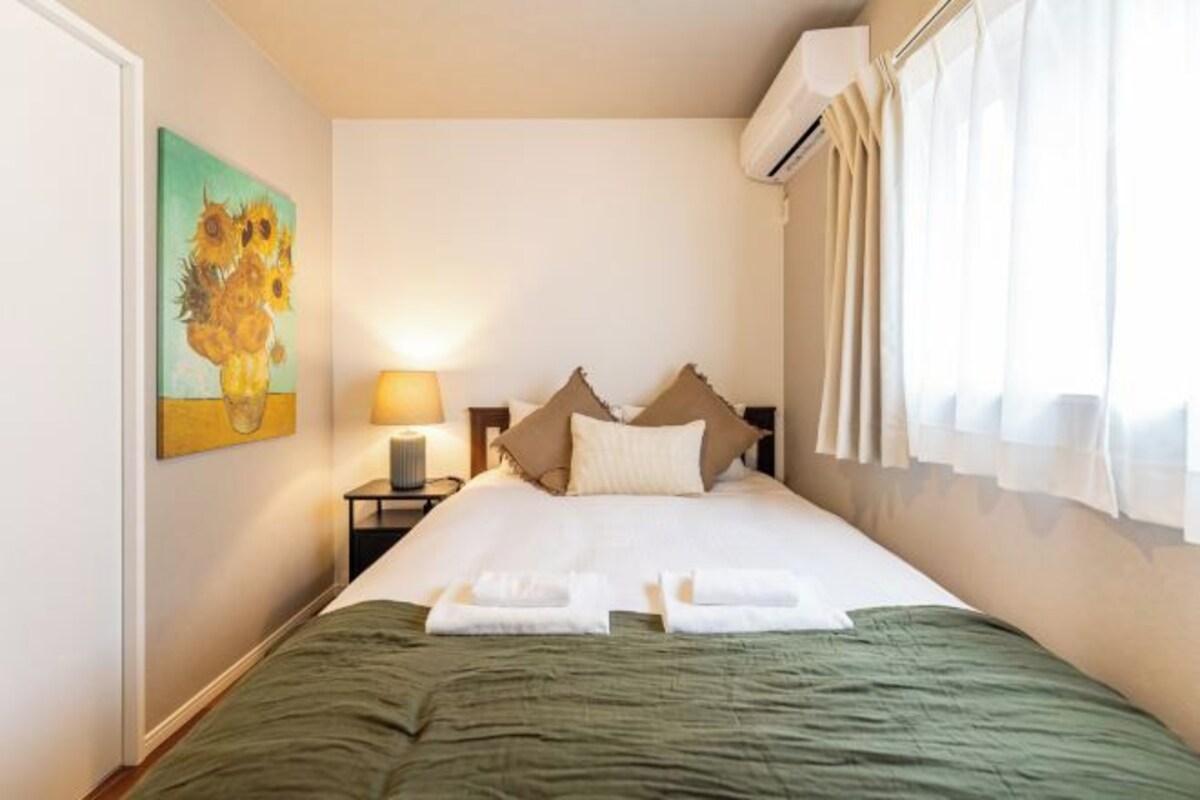 Quarto de hotel com cama, almofadas, mesa de cabeceira, luminária, ar condicionado e um quadro na parede.