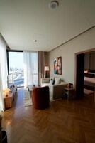 Quarto de hotel moderno com uma janela grande, um sofá, uma mesa de centro e um banquinho vermelho.