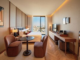 Quarto de hotel com uma cama grande, duas poltronas, uma mesa redonda pequena, uma escrivaninha com televisão e vista para a cidade.