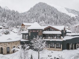 Um edifício grande, com vários andares e uma varanda saliente, rodeado por árvores e montanhas cobertas de neve.