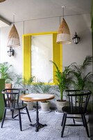 Uma sala de jantar com mesa e cadeiras, uma janela com moldura amarela, luminárias pendentes e plantas em vasos.