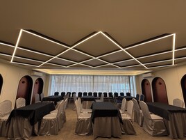 Uma sala de conferências com mesas e cadeiras, iluminação de teto geométrica e amplas janelas com cortinas.