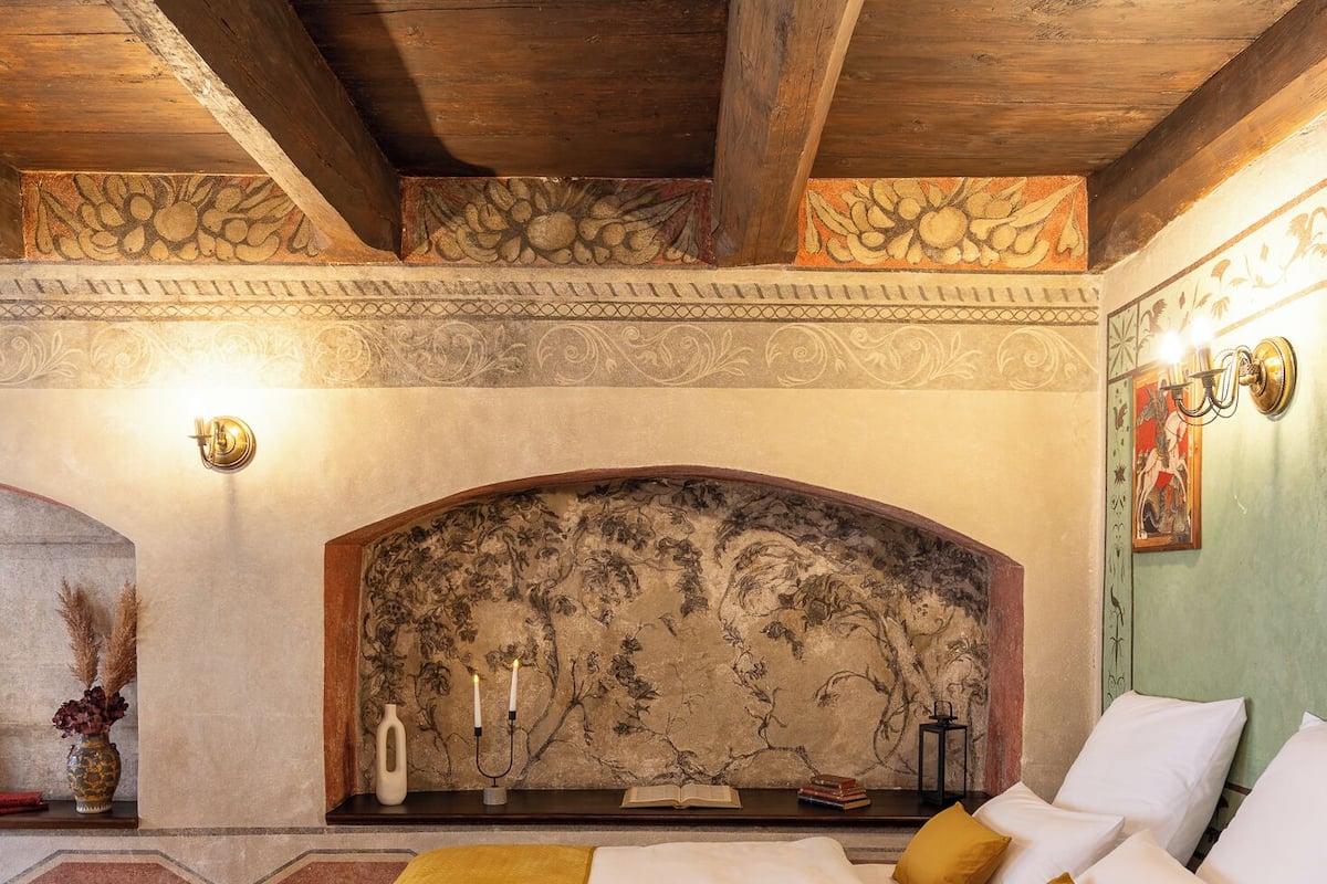 Quarto com uma cama grande, vigas de madeira, lareira decorativa e mesa de cabeceira com velas.