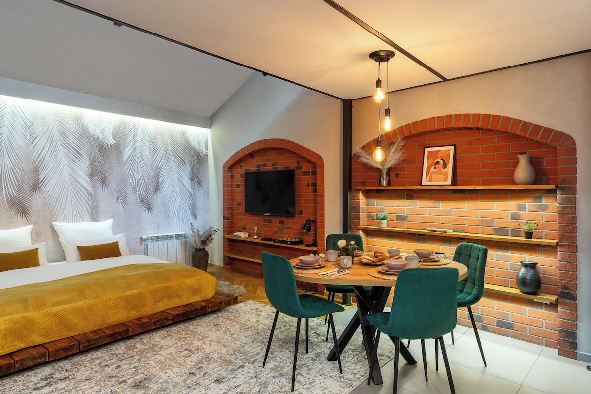 Quarto moderno com cama grande, televisão fixada em parede de tijolos e área de refeições com cadeiras verdes e uma mesa.