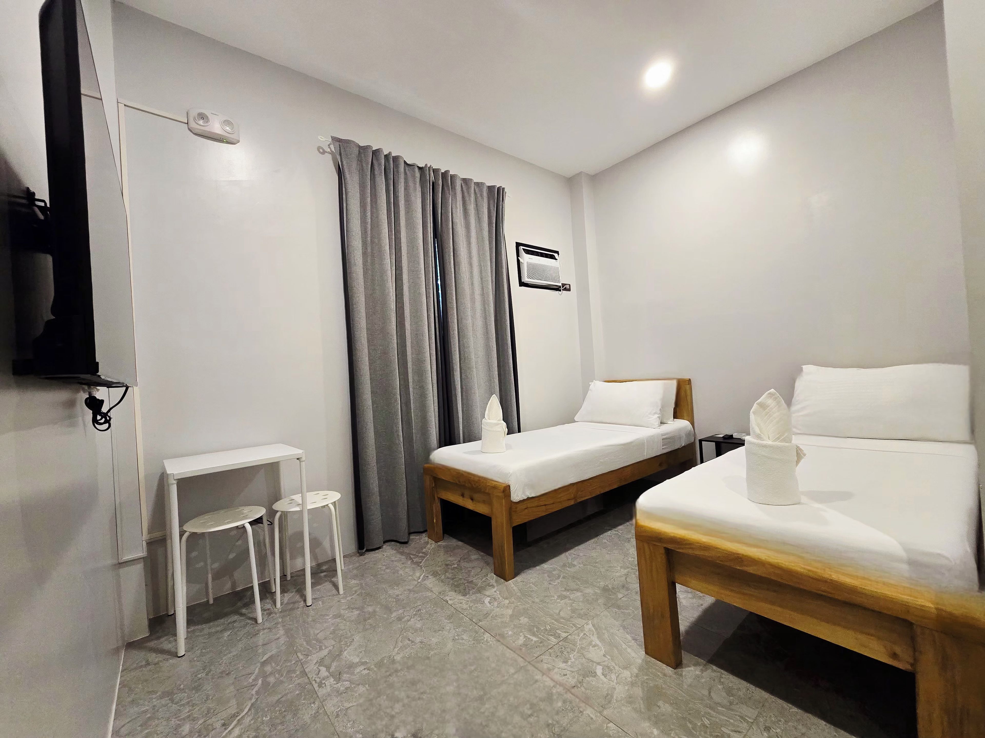 Quarto de hotel com duas camas de solteiro, uma mesa pequena, duas cadeiras, uma TV fixada na parede e uma janela com cortinas.