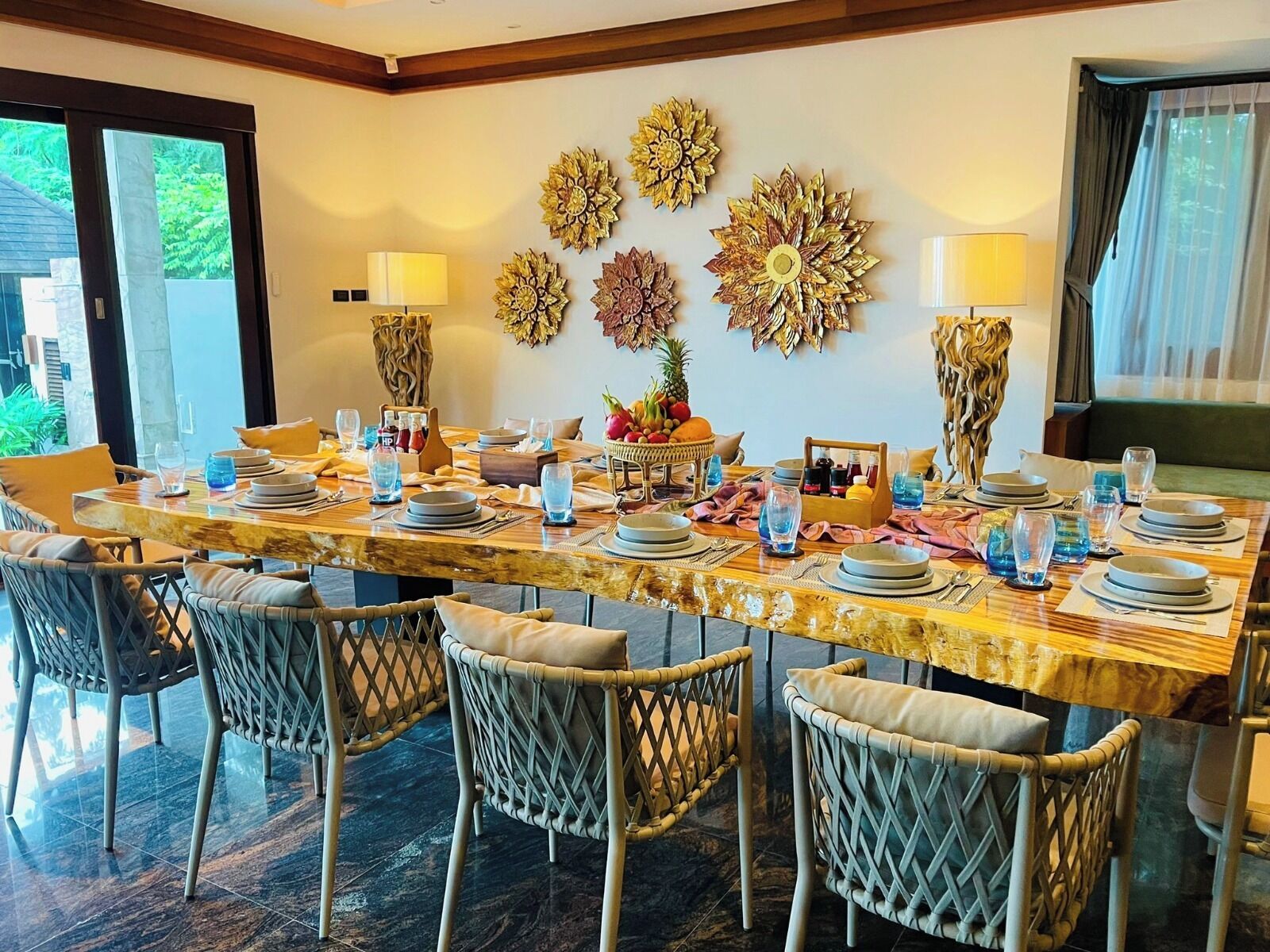 Uma área de refeições com uma mesa comprida posta para vários convidados, cercada por cadeiras de vime.