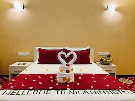 Quarto de hotel com cama, colcha vermelha e branca, decoração de toalha em formato de coração, cesta de frutas e placa de boas-vindas.