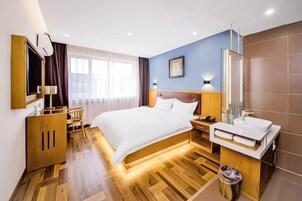 Quarto de hotel com cama, mesa de cabeceira, luminária, espelho e cômoda de madeira.
