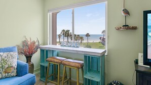 Quarto com vista para a praia, área de bar com banquetas, sofá azul e um pássaro decorativo na parede.