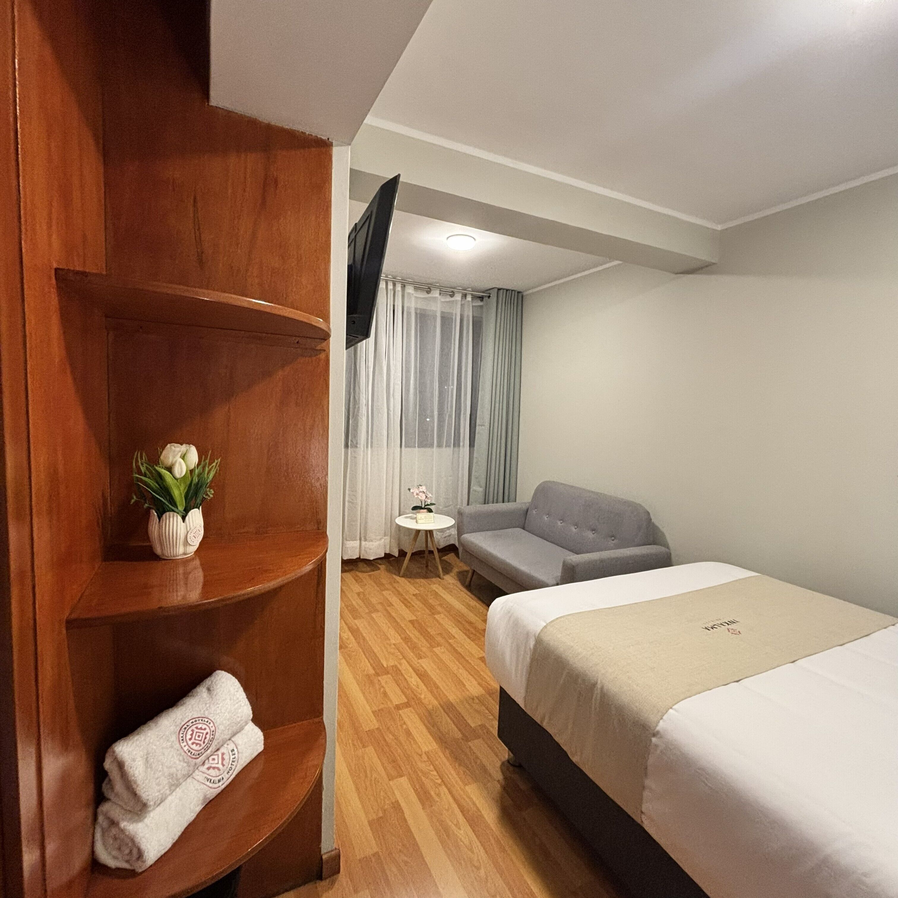 Quarto de hotel com prateleira de madeira, sofá cinza, cama com colcha branca e pequena mesa com um vaso de flores.