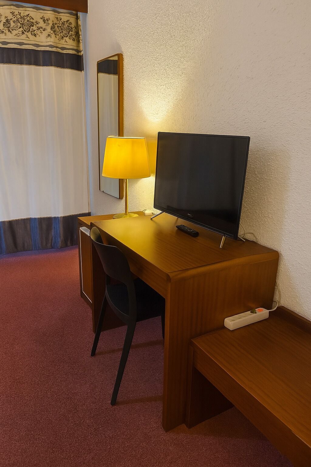 Quarto de hotel com escrivaninha de madeira, uma cadeira preta, televisão, luminária e mesa de cabeceira.