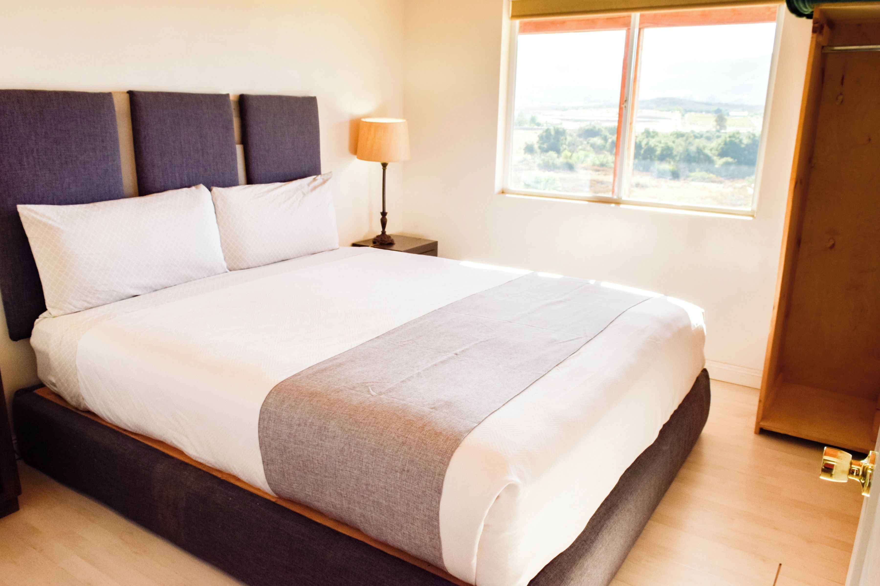 Cama bem arrumada com cabeceira, luminária de cabeceira e janela com vista para o exterior.