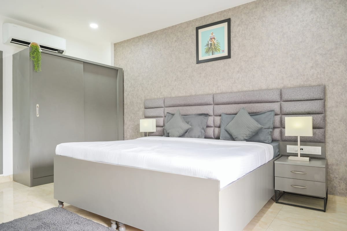 Quarto moderno com cama grande, cabeceira cinza, mesas de cabeceira com abajures e ar-condicionado fixado na parede.