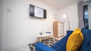Uma sala de estar moderna com um sofá azul, uma mesinha branca com uma planta em vaso, um armário de madeira e uma TV na parede exibindo um logotipo.