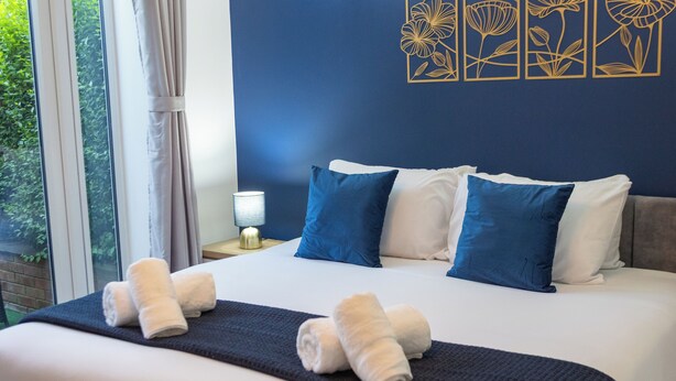 Cama bem arrumada com almofadas brancas e azuis, um cobre-leito azul escuro e duas toalhas enroladas na cama.