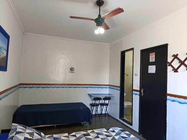 Quarto pequeno com uma cama, escrivaninha, duas cadeiras, ventoinha no teto e uma porta com uma placa.
