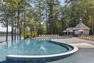 Piscina com borda curva, rodeada por espreguiçadeiras e um pequeno edifício com telhado em azulejos.