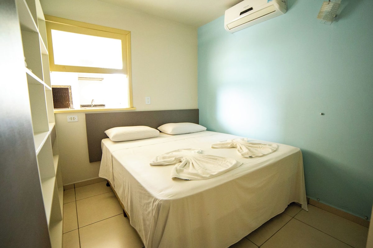 Quarto pequeno e bem arrumado, com cama, almofadas e janela.