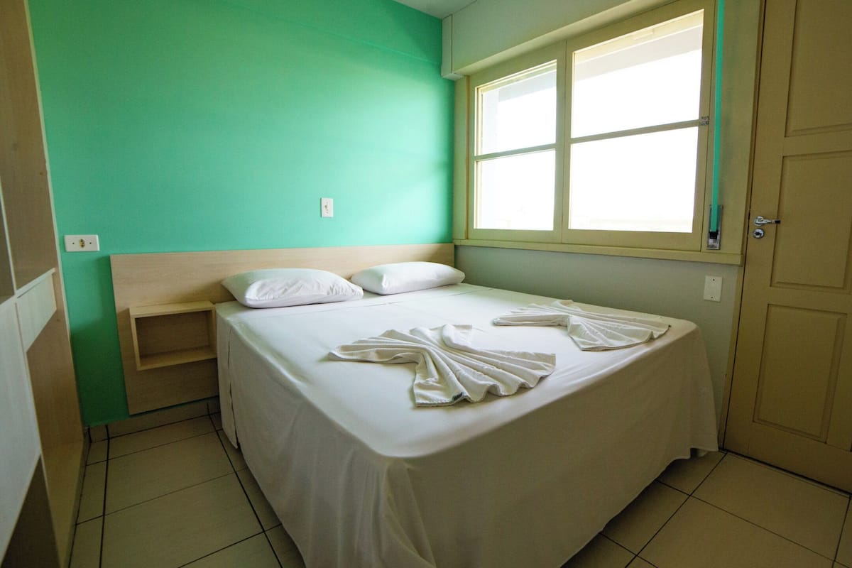 Uma cama de solteiro com roupa de cama branca, cabeceira de madeira e janela com persianas.