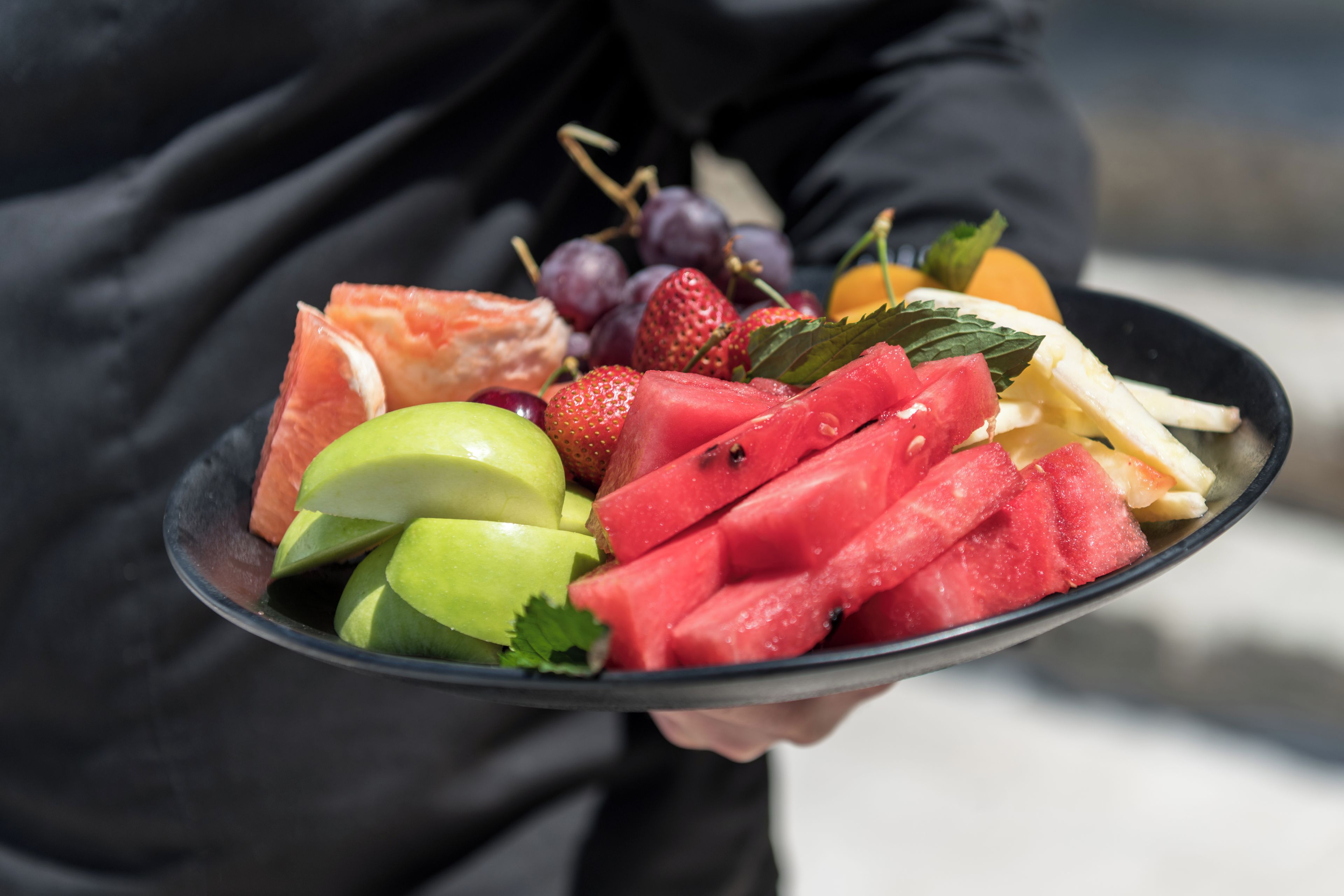 Uma pessoa segurando uma tigela com frutas variadas, incluindo melancia, morangos, uvas e maçãs.