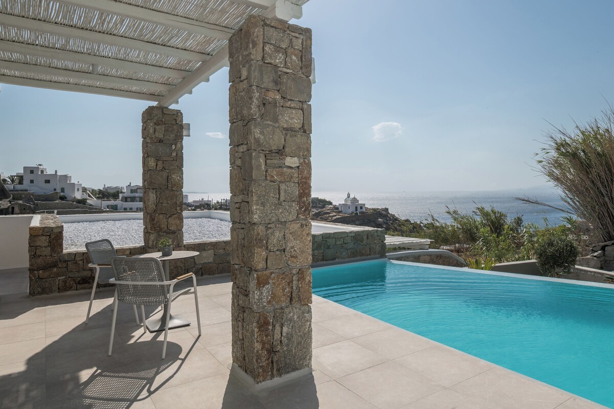 Uma área à beira da piscina com pilares de pedra, um espaço coberto para sentar e vista para o mar.