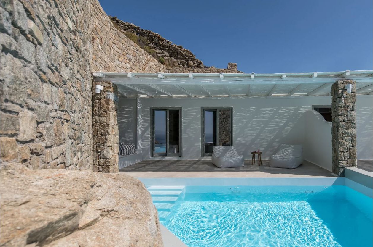 Área de piscina com paredes de pedra, piscina azul límpida, pilares de pedra e área de estar coberta.
