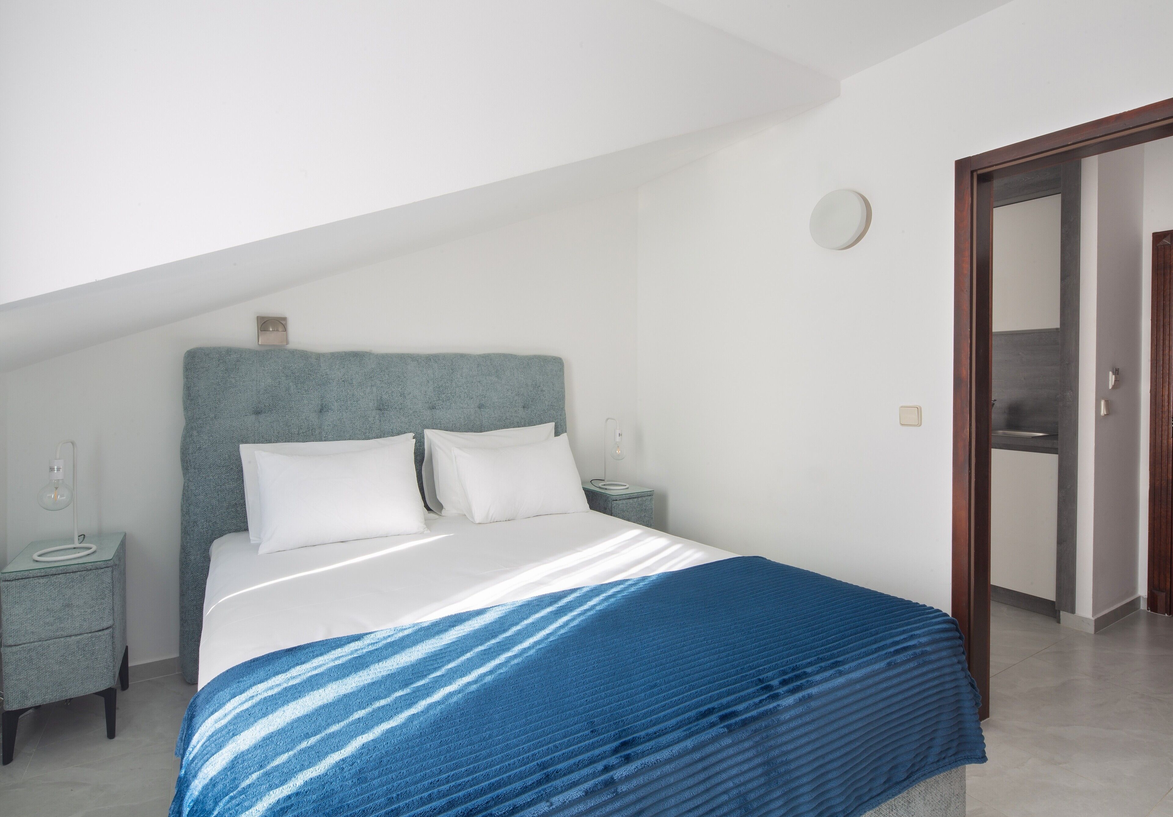 Quarto com cama, mesa de cabeceira, relógio na parede e porta para um banheiro.