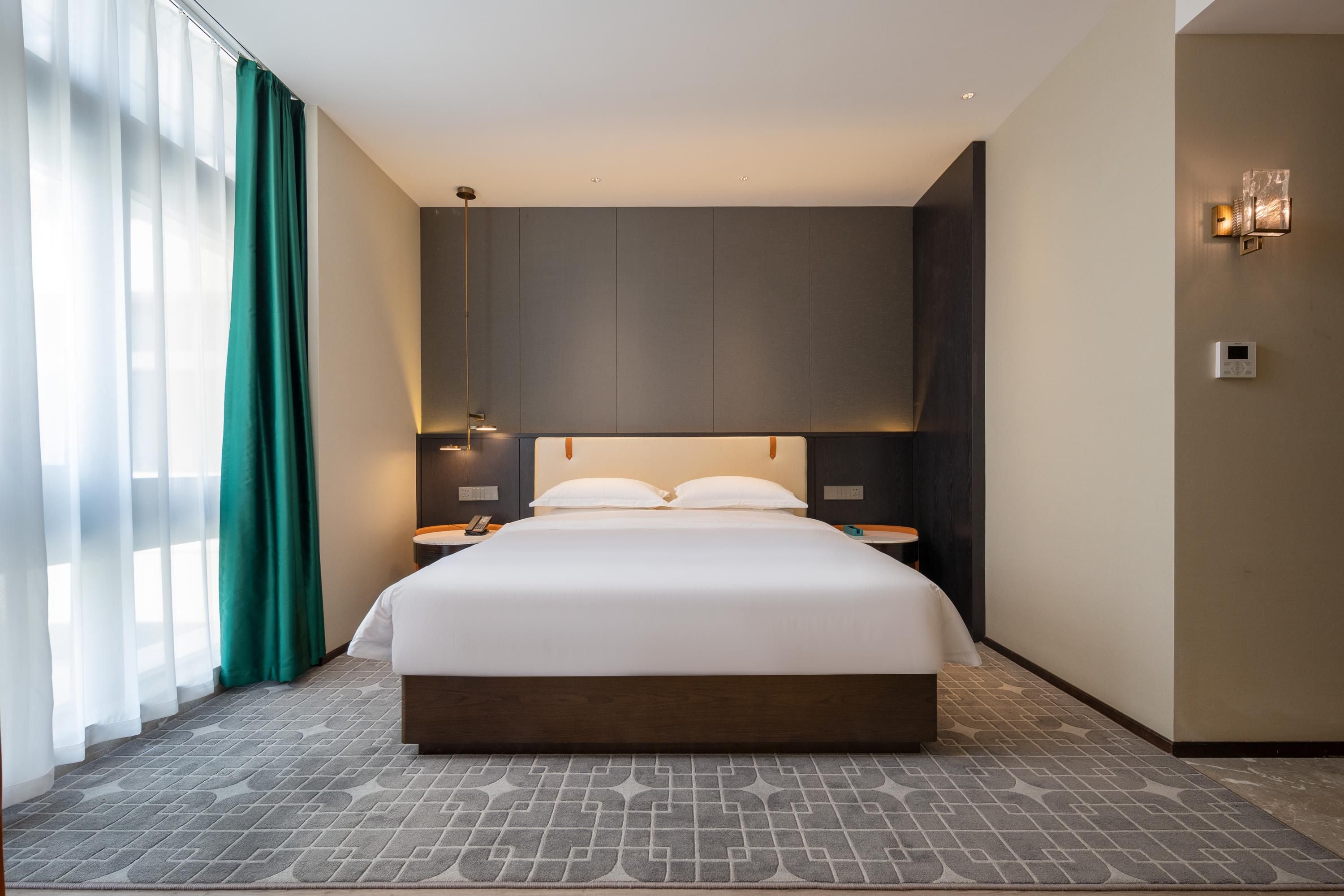 Quarto de hotel moderno com uma cama grande, mesa de cabeceira, abajur e vista da cidade pela janela.