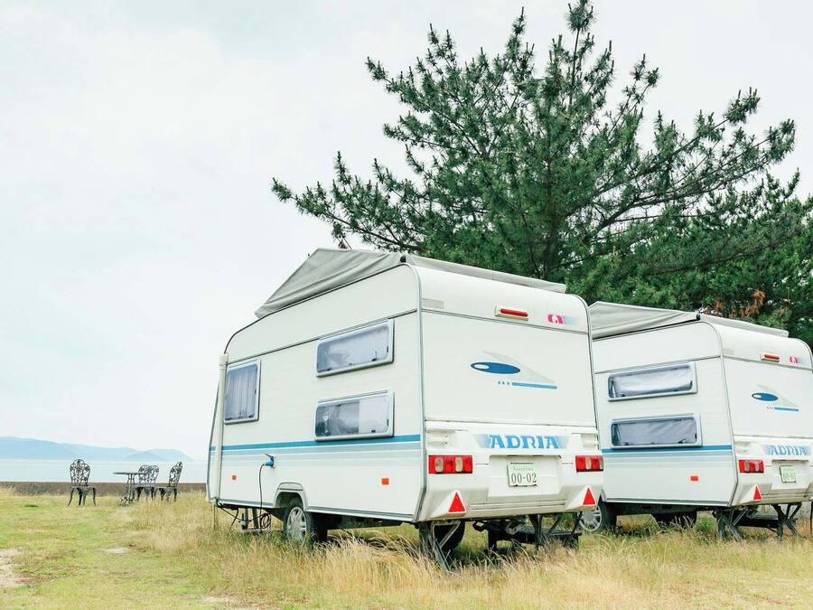 Duas RVs brancas com a marca 'ADRIA' estacionadas em um campo gramado, com uma grande árvore e um corpo d'água ao fundo.