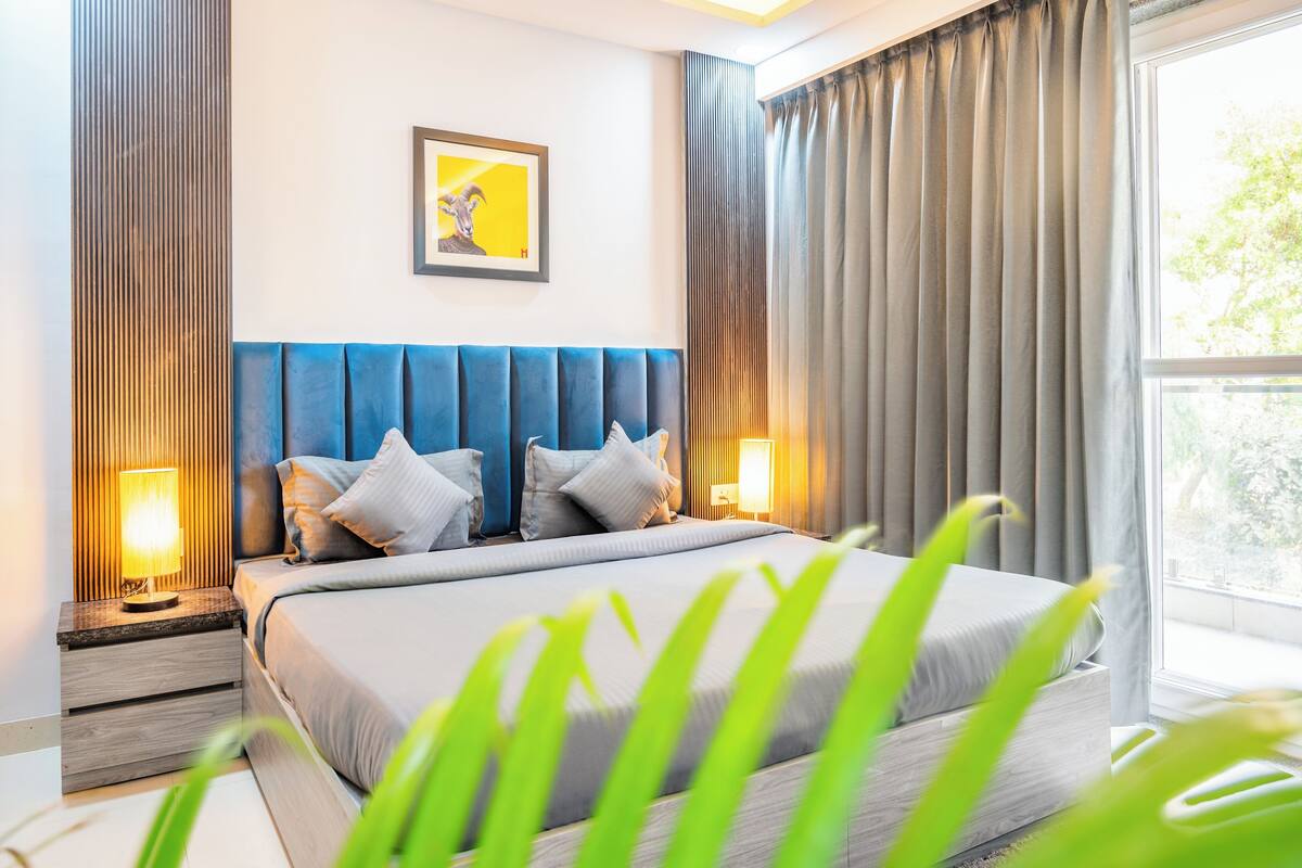 Quarto moderno com cama grande, duas luminárias de cabeceira, uma obra de arte emoldurada na parede e vista para o verde pela janela.