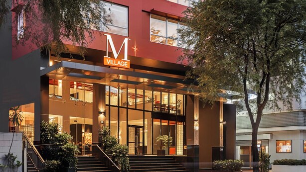 Um hotel de vários andares com um logotipo 'M' em destaque e a placa 'VILLAGE', apresentando uma fachada moderna, grandes janelas e uma entrada bem iluminada.