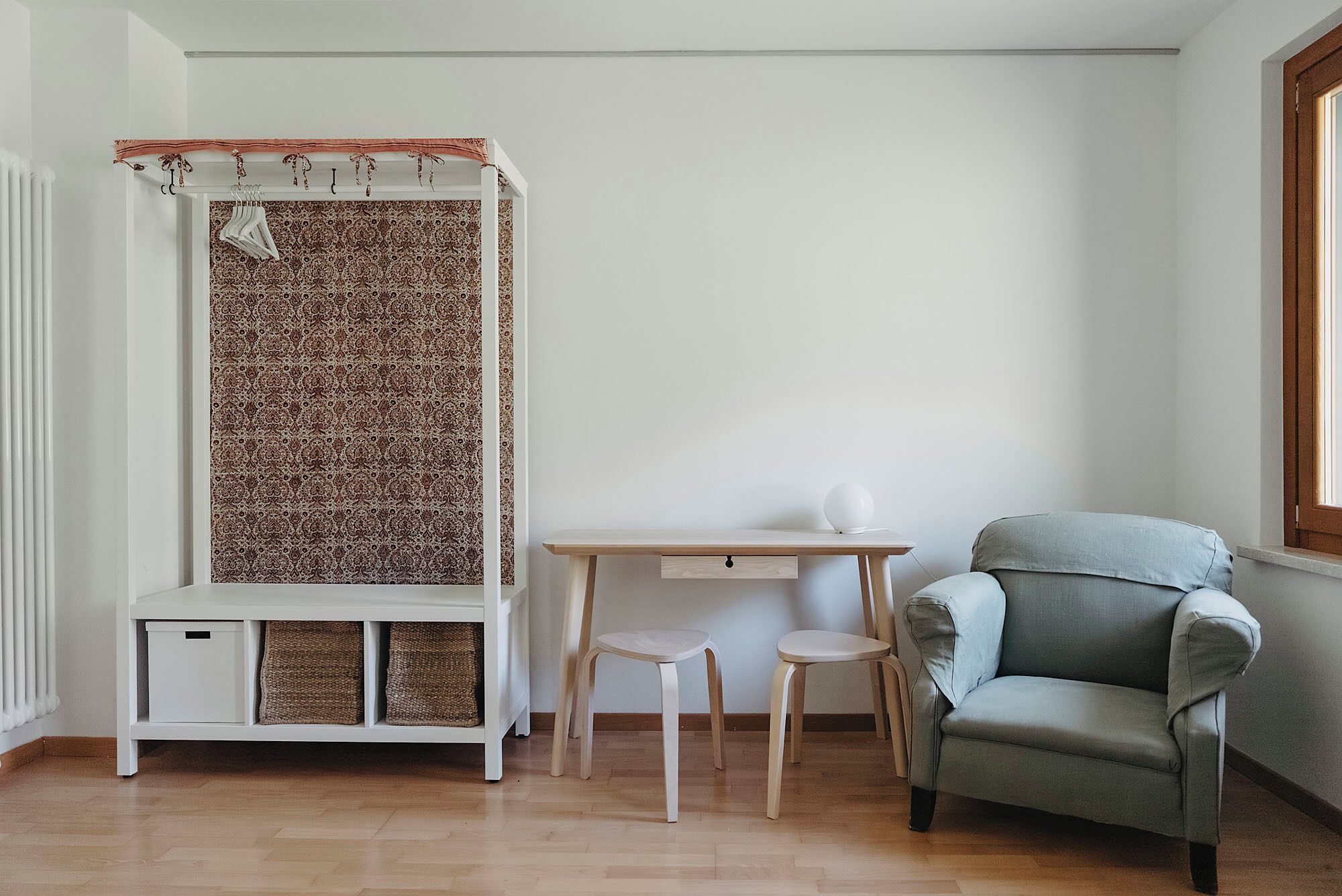 Quarto com guarda-roupa branco, uma pequena escrivaninha, duas cadeiras e uma janela.
