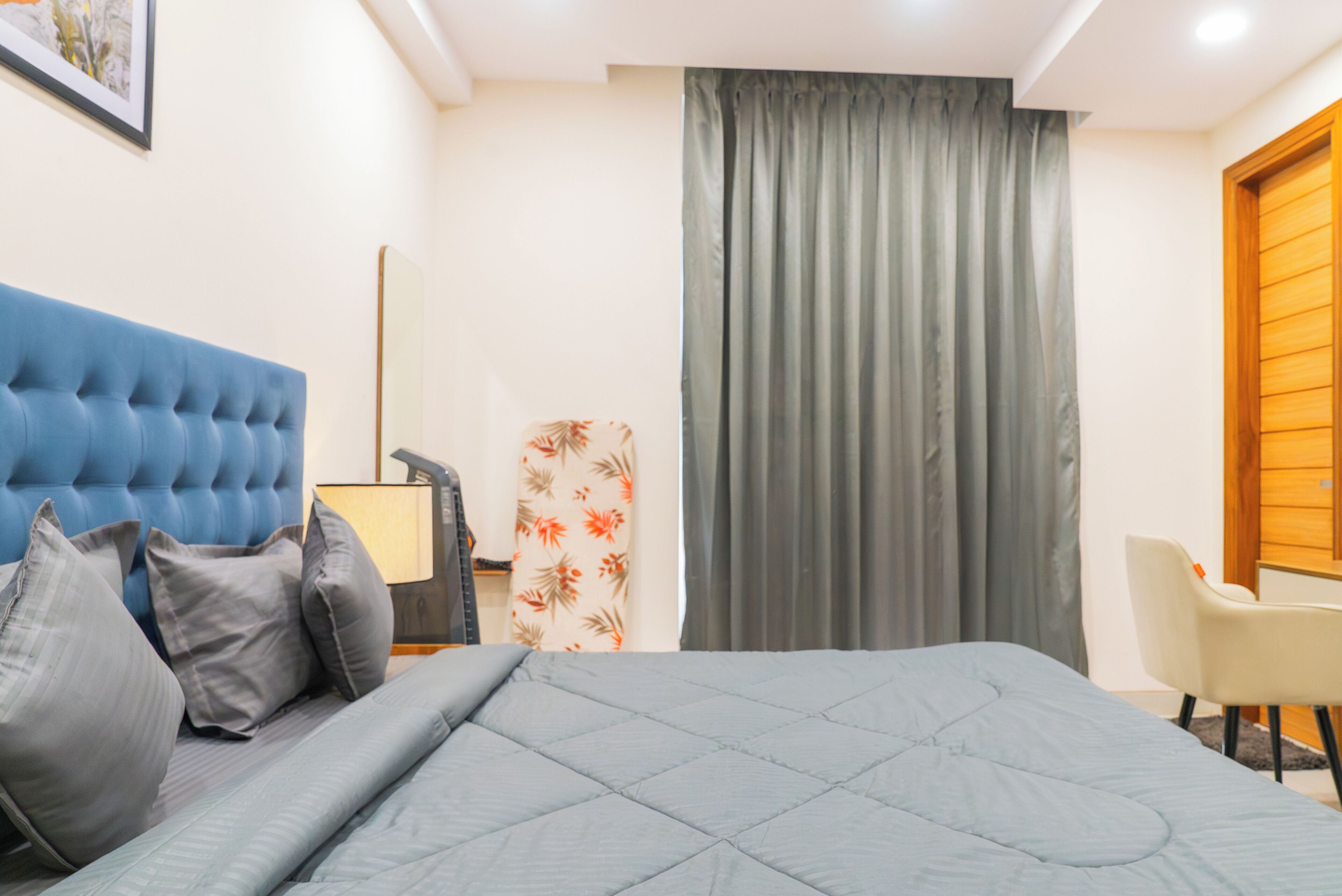 Um quarto com cabeceira azul, cama com roupa de cama cinza, uma cadeira, uma escrivaninha com abajur e uma cortina cinza grande.