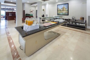 Um buffet de café da manhã com diversas opções de alimentos e bebidas, um balcão de vidro e uma área de cozinha ao fundo.