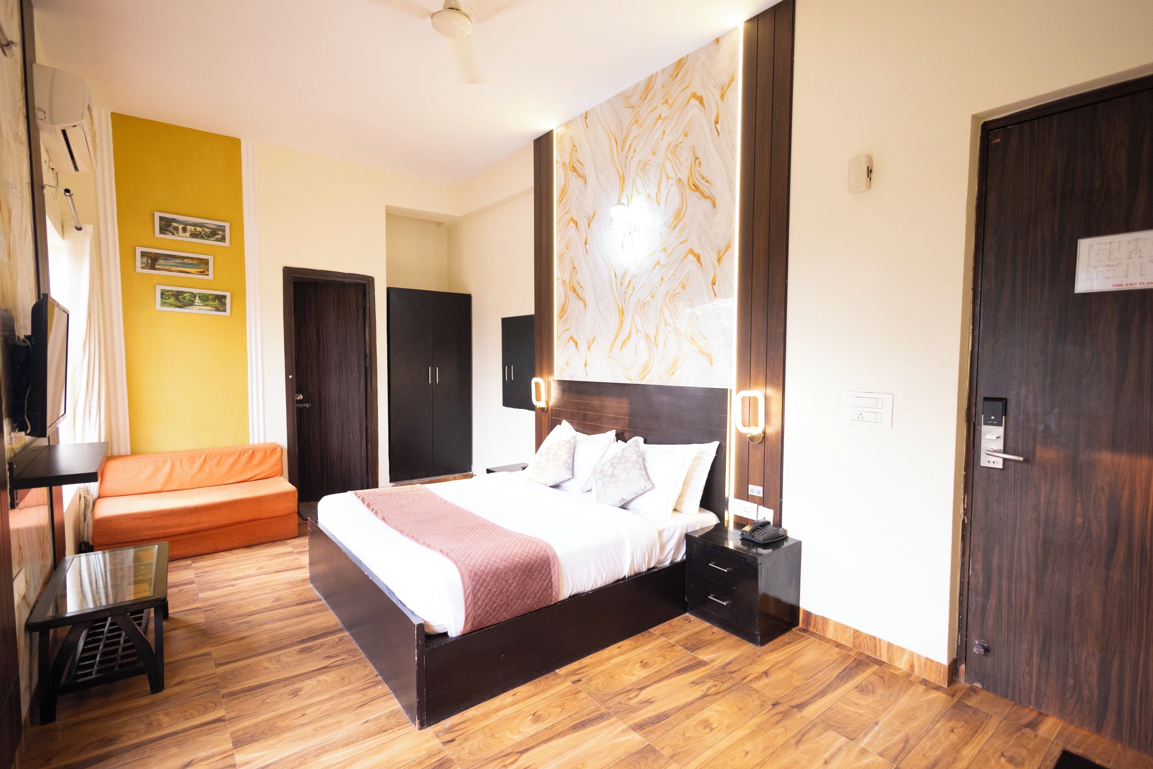 Quarto de hotel moderno com piso de madeira, uma cama grande com roupa de cama branca, uma mesinha de cabeceira e um aplique na parede.