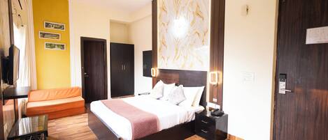 Quarto de hotel moderno com piso de madeira, uma cama grande com roupa de cama branca, uma mesinha de cabeceira e um aplique na parede.