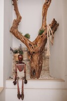 Uma escultura rústica de madeira com uma corda, uma cesta de vime com plantas e um pequeno vaso com planta numa estante.