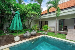 Uma área à beira da piscina com deck de madeira, guarda-sóis verdes e plantas em vasos.
