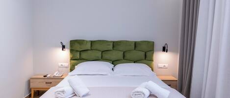 Cama bem arrumada com cabeceira estofada verde, lençóis brancos e duas mesas de cabeceira com luminárias.
