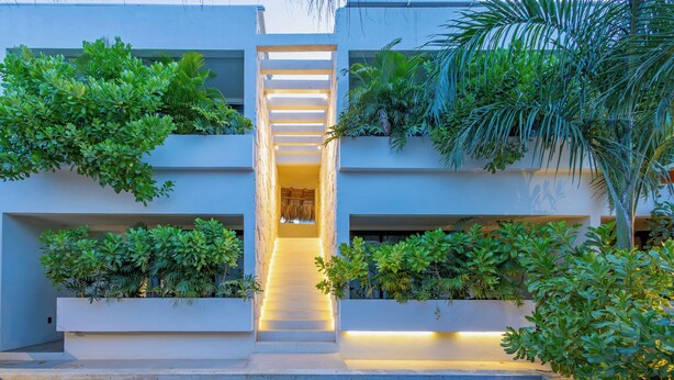 Um edifício moderno com piscina, escadaria que leva a uma entrada coberta e vegetação exuberante ao redor.