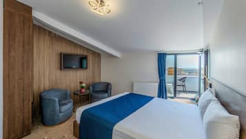 Quarto de hotel com cama, dois poltronas, uma mesinha com uma planta, televisão na parede e varanda com mesa e cadeiras.