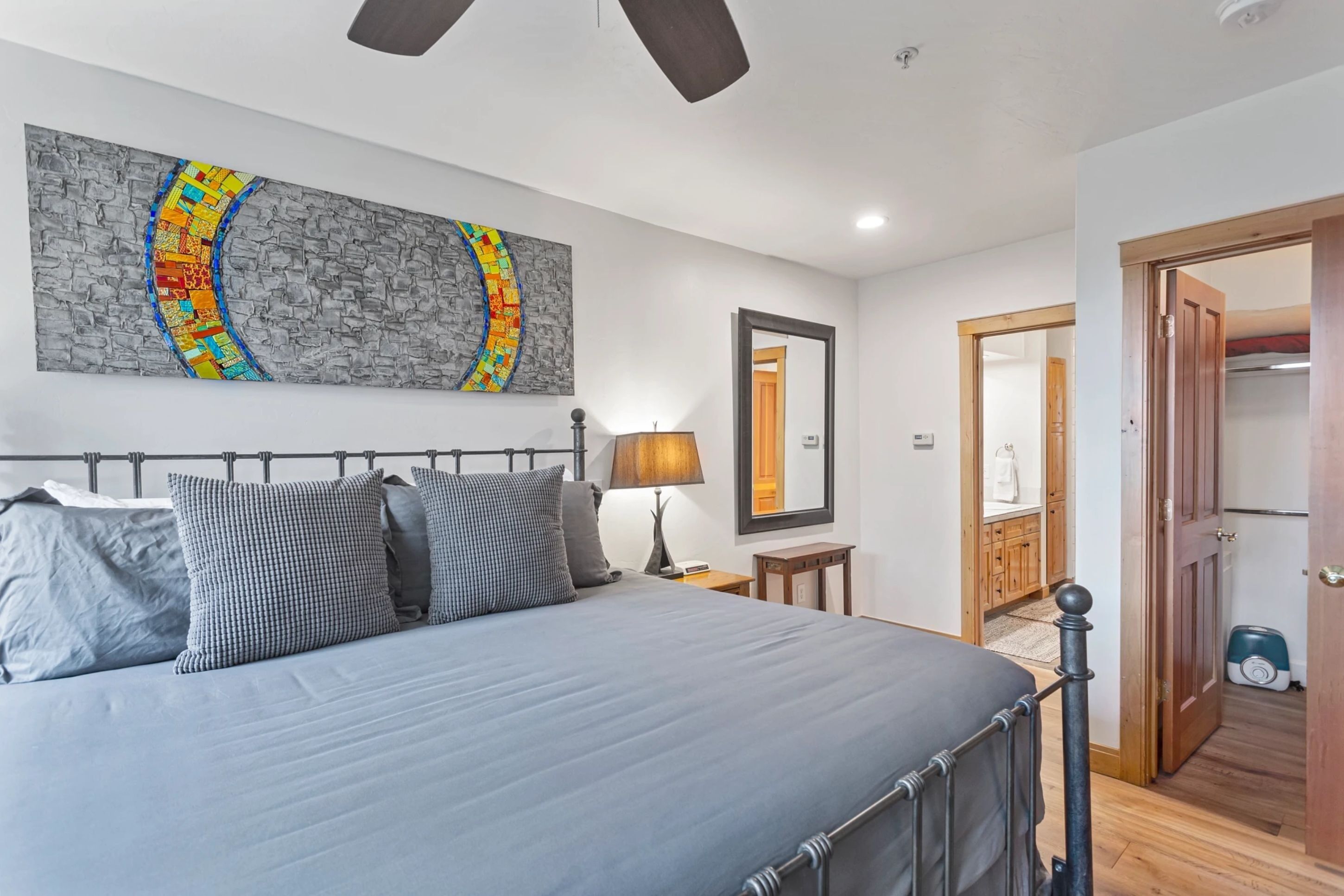 Quarto com cama grande, ventoinha no teto, obra de arte na parede, mesa de cabeceira com abajur e porta de madeira que dá para outro cômodo.