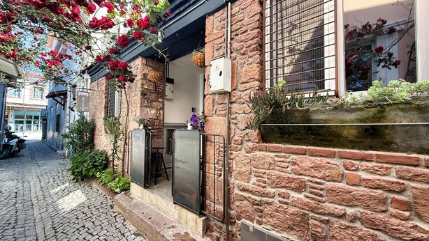 Uma rua estreita de paralelepípedos com um edifício de tijolos, com uma pequena área de estar ao ar livre com cadeiras e uma mesa de metal, uma janela com grade de metal e um cesto suspenso com flores vermelhas.