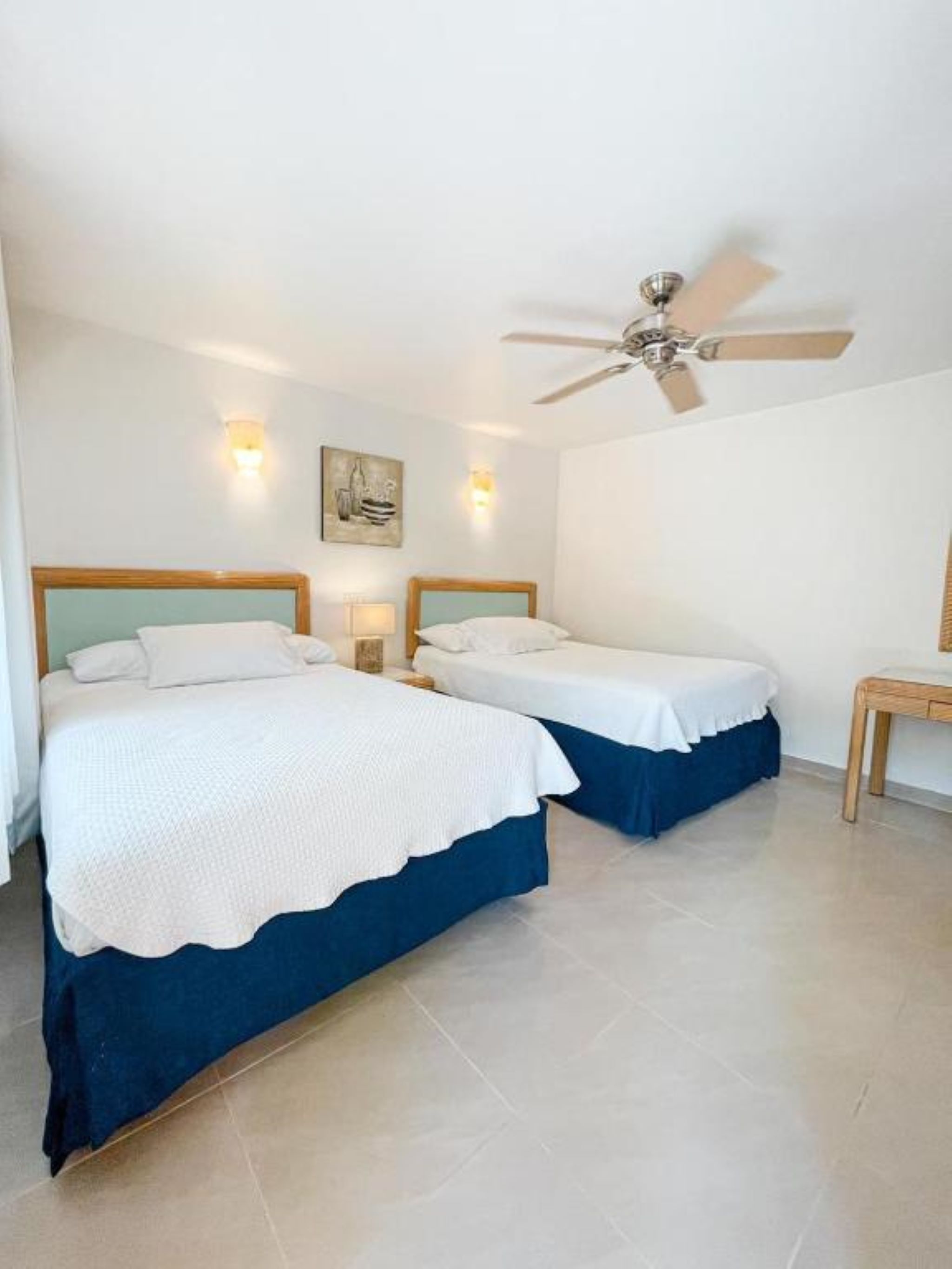 Quarto de hotel com duas camas, um ventilador de teto, luminárias fixadas na parede e uma pequena mesa de madeira.