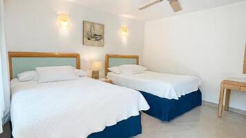 Quarto de hotel com duas camas, um ventilador de teto, luminárias fixadas na parede e uma pequena mesa de madeira.